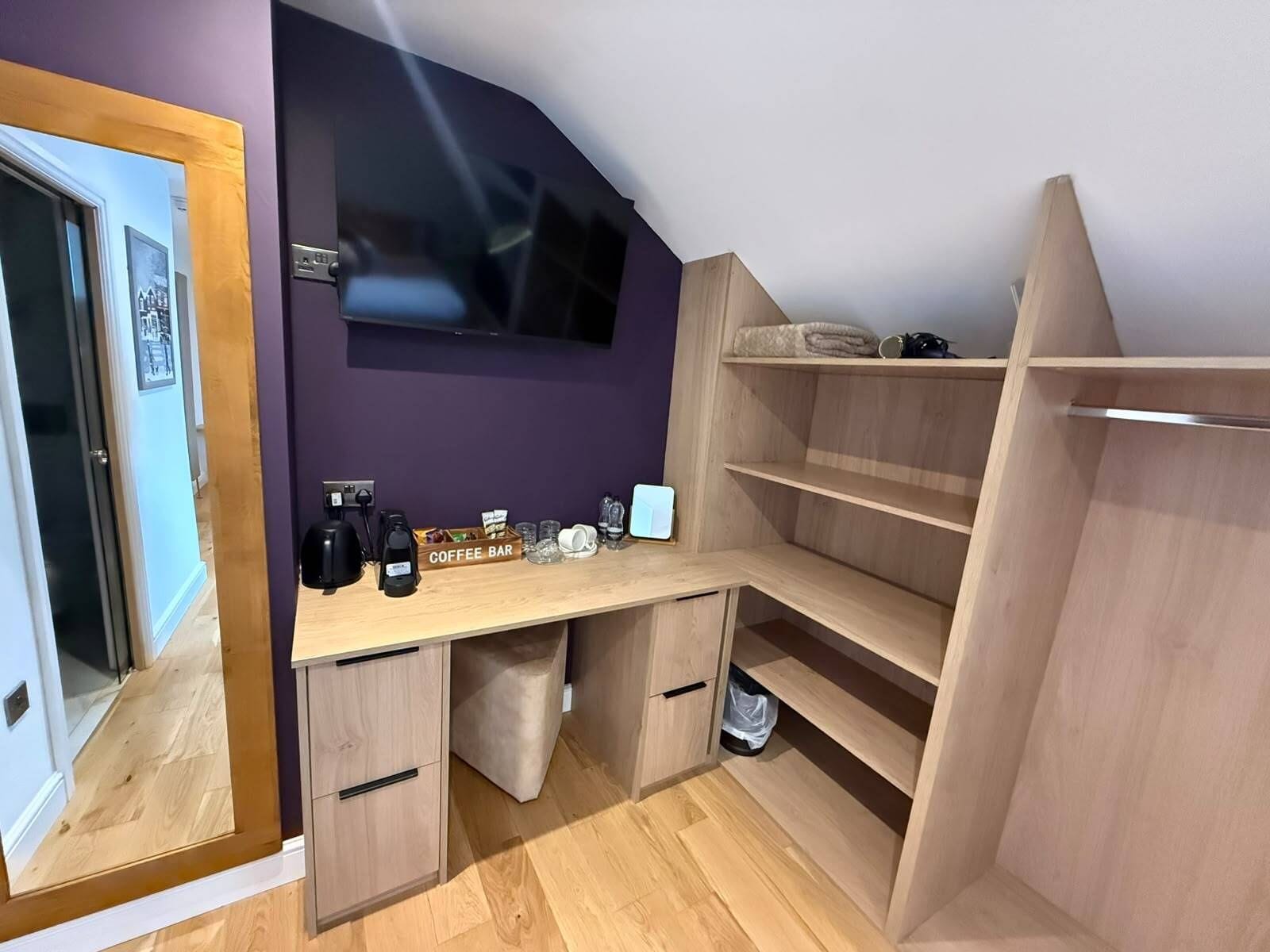 Quarto compacto com escrivaninha, espaço para café e TV fixada na parede.