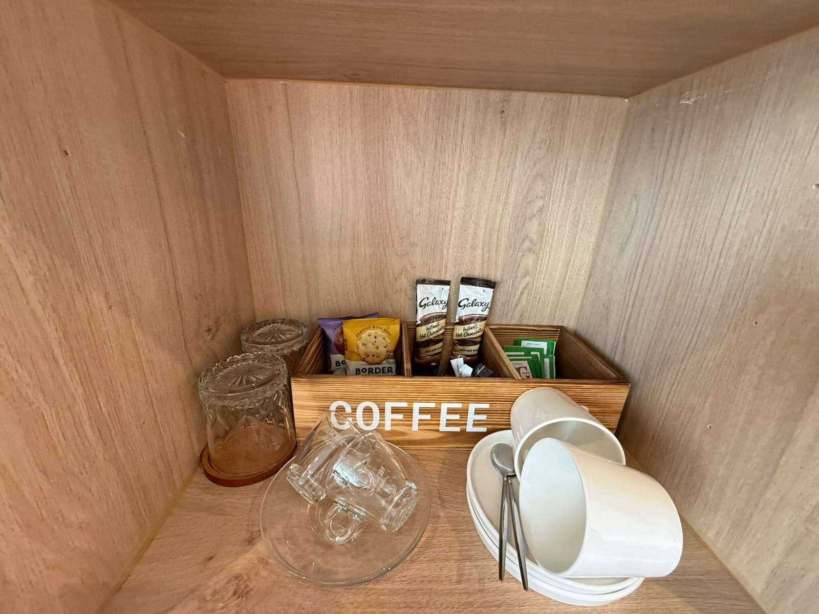 Caixa de madeira com a etiqueta 'COFFEE' contendo embalagens de café, um copo e um saquinho de chá.