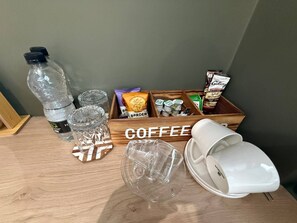 Uma mesa de madeira com um espaço para café montado, incluindo uma cafeteira, chávenas, uma garrafa de água, copos e uma caixa com a etiqueta 'COFFEE' contendo diversos produtos de café.