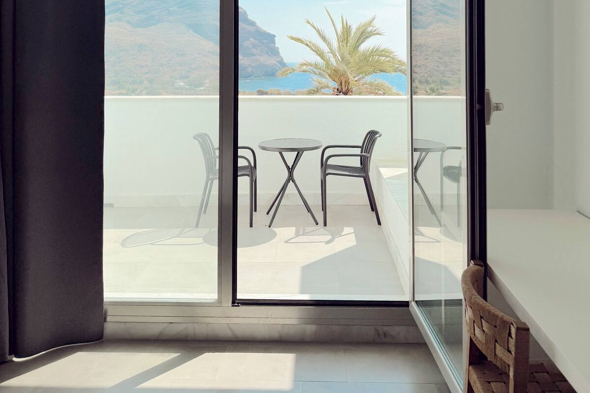 Varanda com mesa e cadeiras, vista para o mar e montanhas, e porta de vidro deslizante.