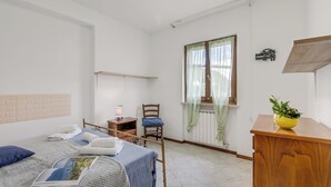 Quarto com cama, cadeira, mesa de cabeceira, janela com cortinas e uma pequena planta em um armário de madeira.