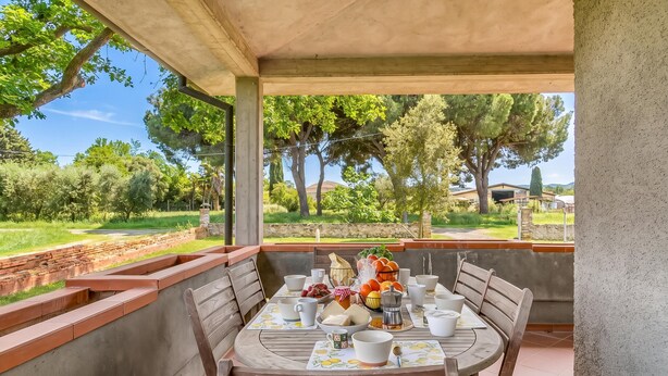 Uma área de refeições coberta ao ar livre, com uma mesa posta para dois, rodeada de cadeiras, com vista para um jardim com árvores e um céu limpo.