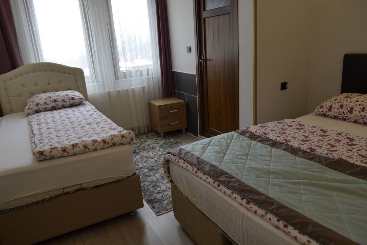 Quarto de hotel com duas camas, uma mesa de cabeceira de madeira, um radiador e uma porta.