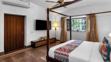 Uma cama de dossel com colcha estampada, um ventilador de teto, uma televisão de tela plana e uma janela com cortinas.