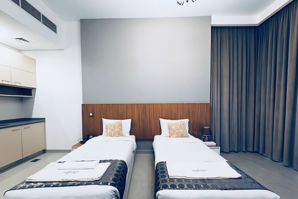 Quarto de hotel com duas camas, uma cozinha compacta, uma cabeceira de madeira e uma janela grande com cortinas.