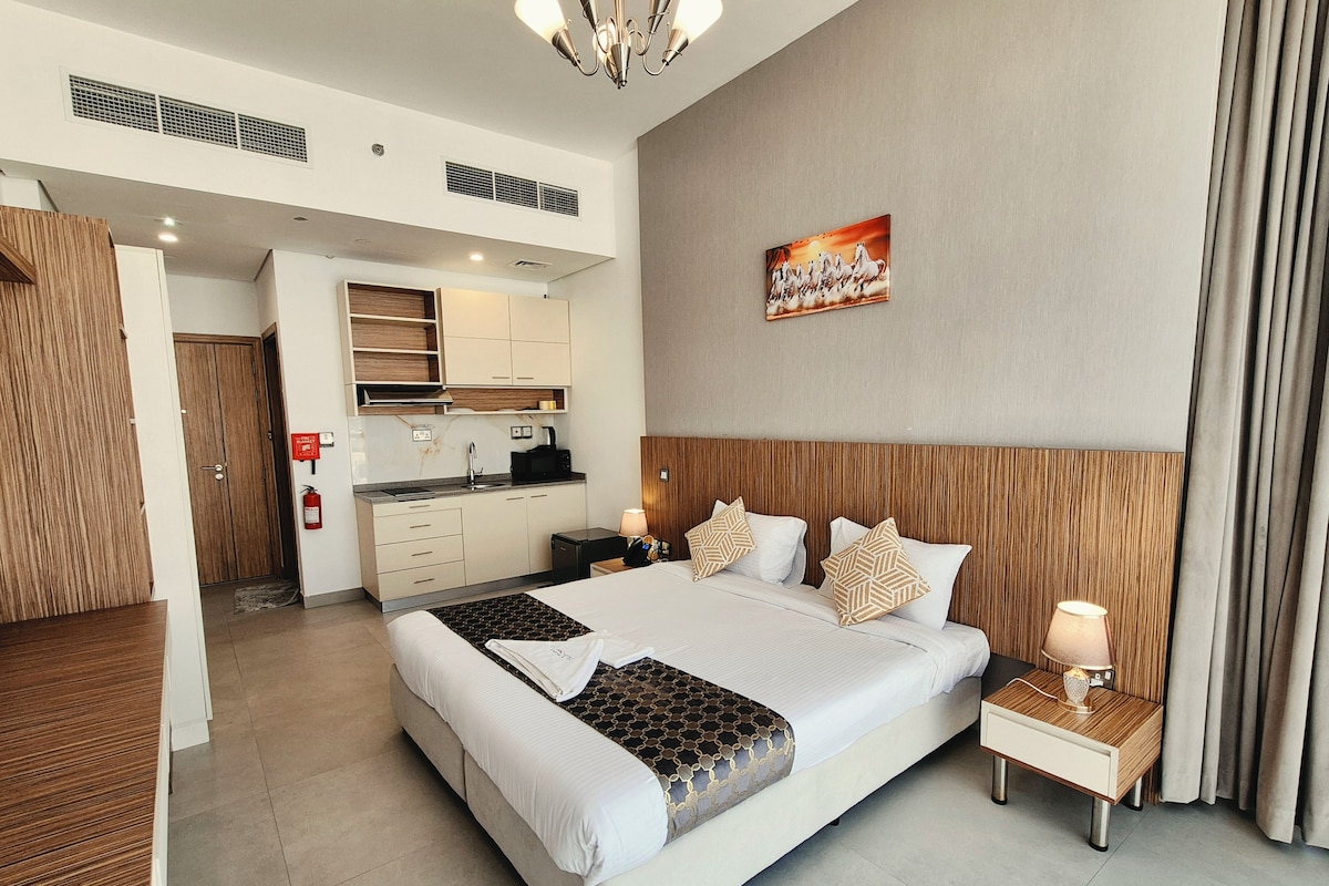 Quarto de hotel com cama grande, mesa de cabeceira com luminária, cabeceira de madeira, cozinha e um quadro na parede.