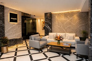 Um lobby moderno com piso xadrez, um conjunto de sofás, uma mesa de centro com arranjo floral e uma placa com a inscrição 'nivasole lara' na parede.