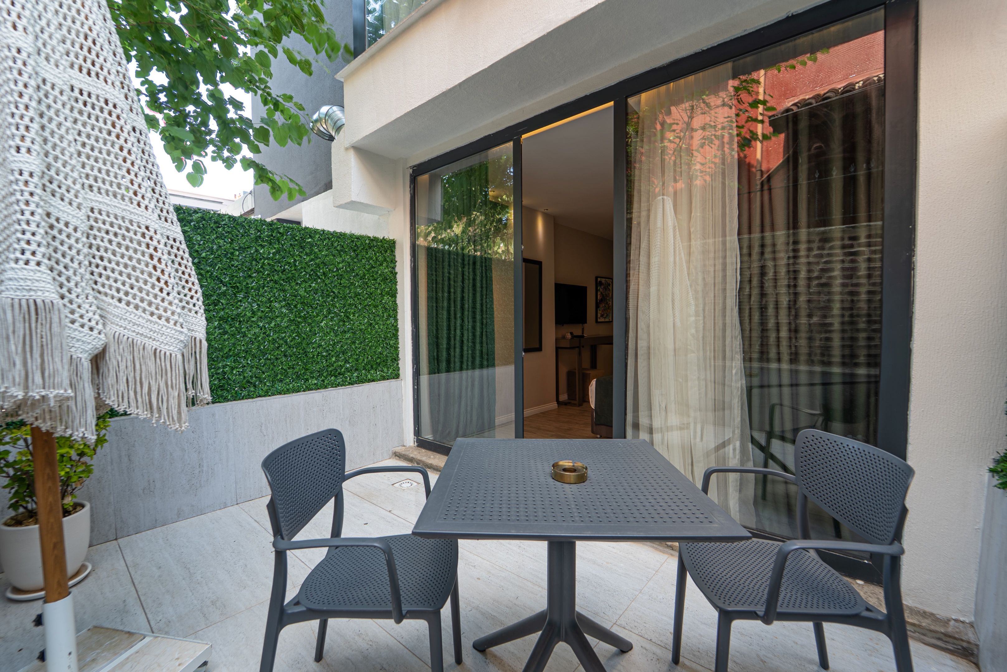 Um pátio externo com mesa e cadeiras, uma parede verde e uma porta de vidro deslizante que dá para um espaço interno.