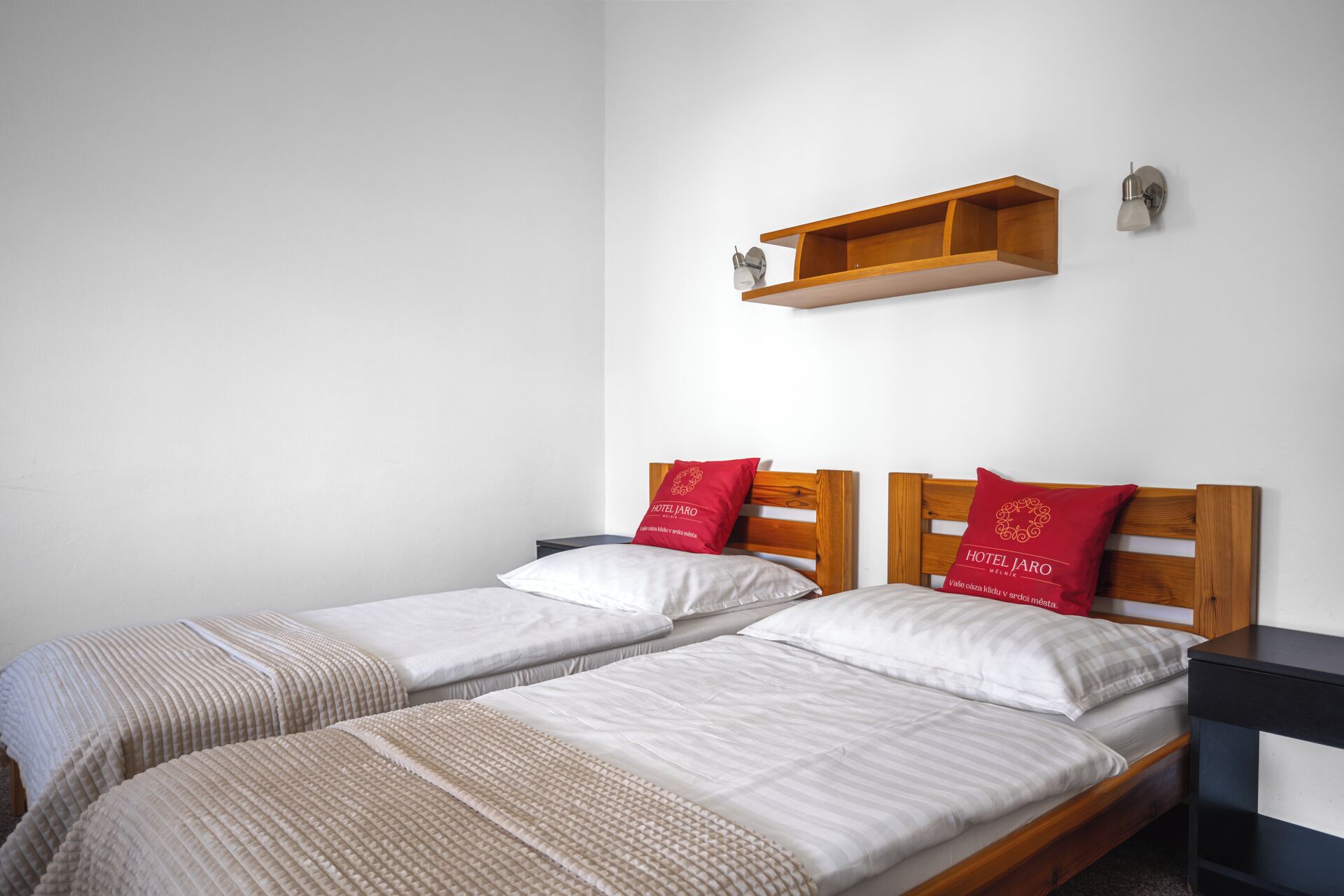Quarto de hotel com duas camas de solteiro, cabeceira de madeira, pequena prateleira sobre as camas e mesa de cabeceira com abajur.