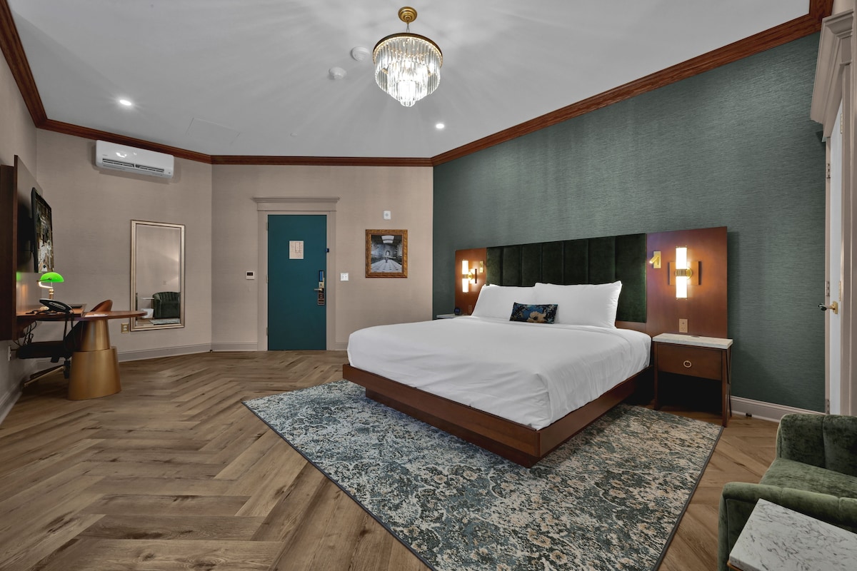 Quarto de hotel com uma cama grande, uma escrivaninha, uma cadeira, luminária de mesa, ar condicionado fixado na parede, lustre e uma porta.