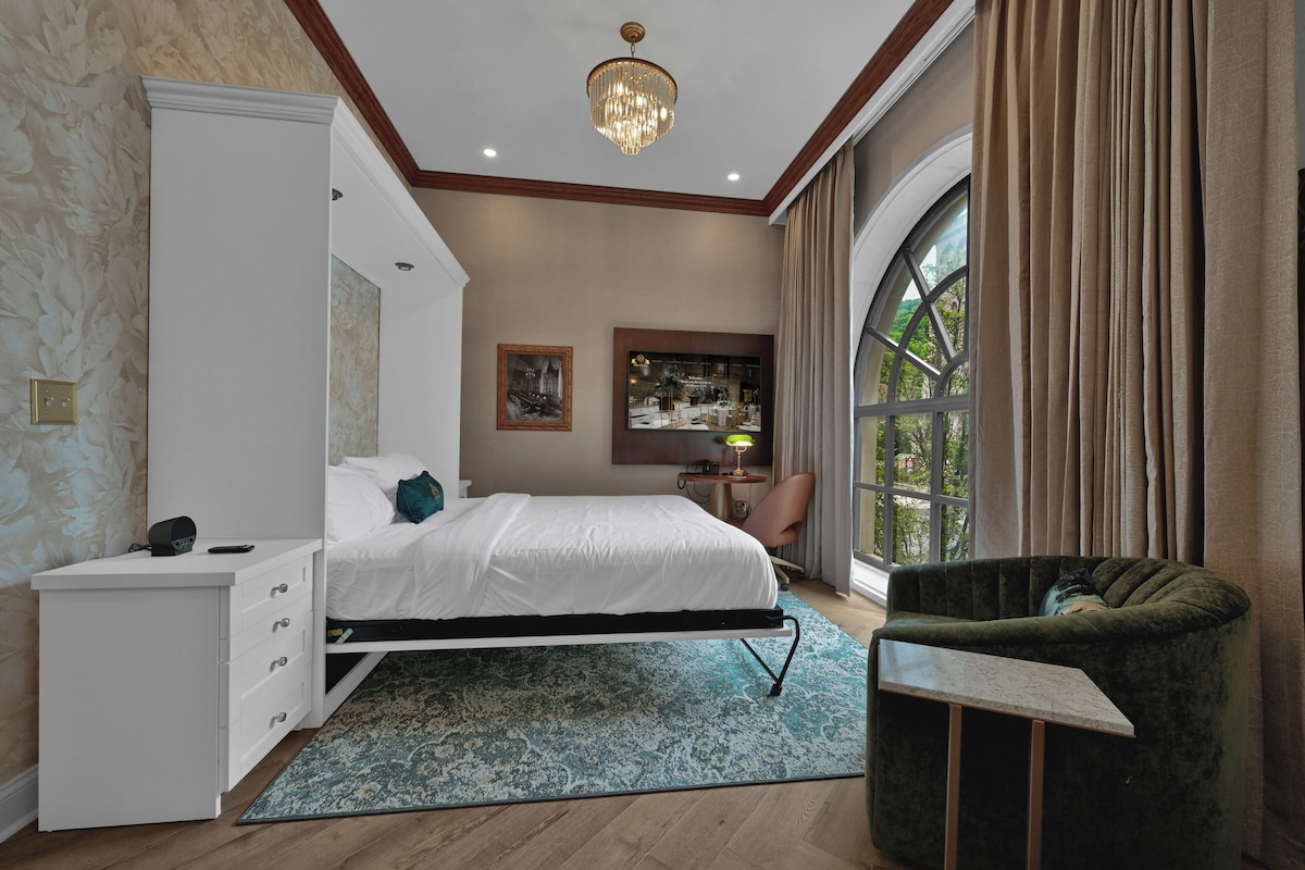 Quarto de hotel com cama, escrivaninha, cadeira, janela ampla e lustre.