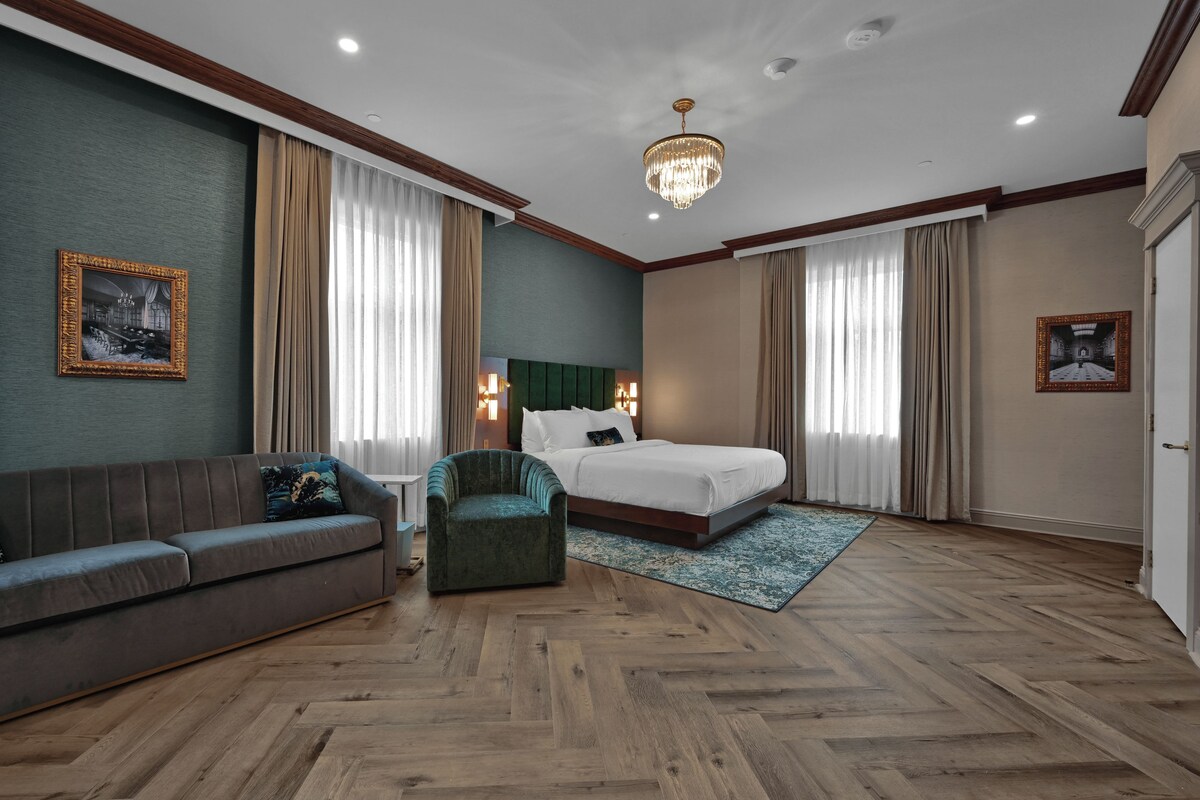 Quarto de hotel com sofá, cadeira, cama, lustre e duas pinturas.