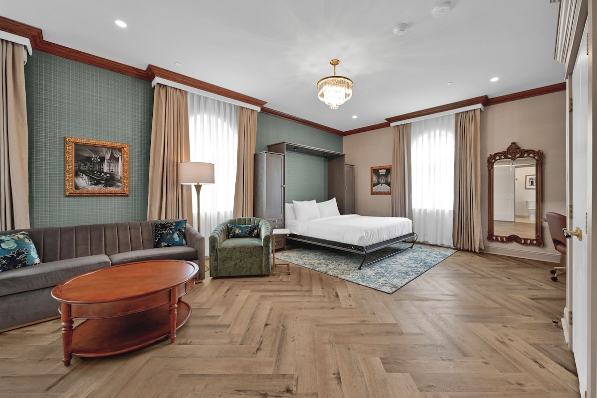 Quarto de hotel com cama, sofá, poltrona e mesa de centro de madeira.