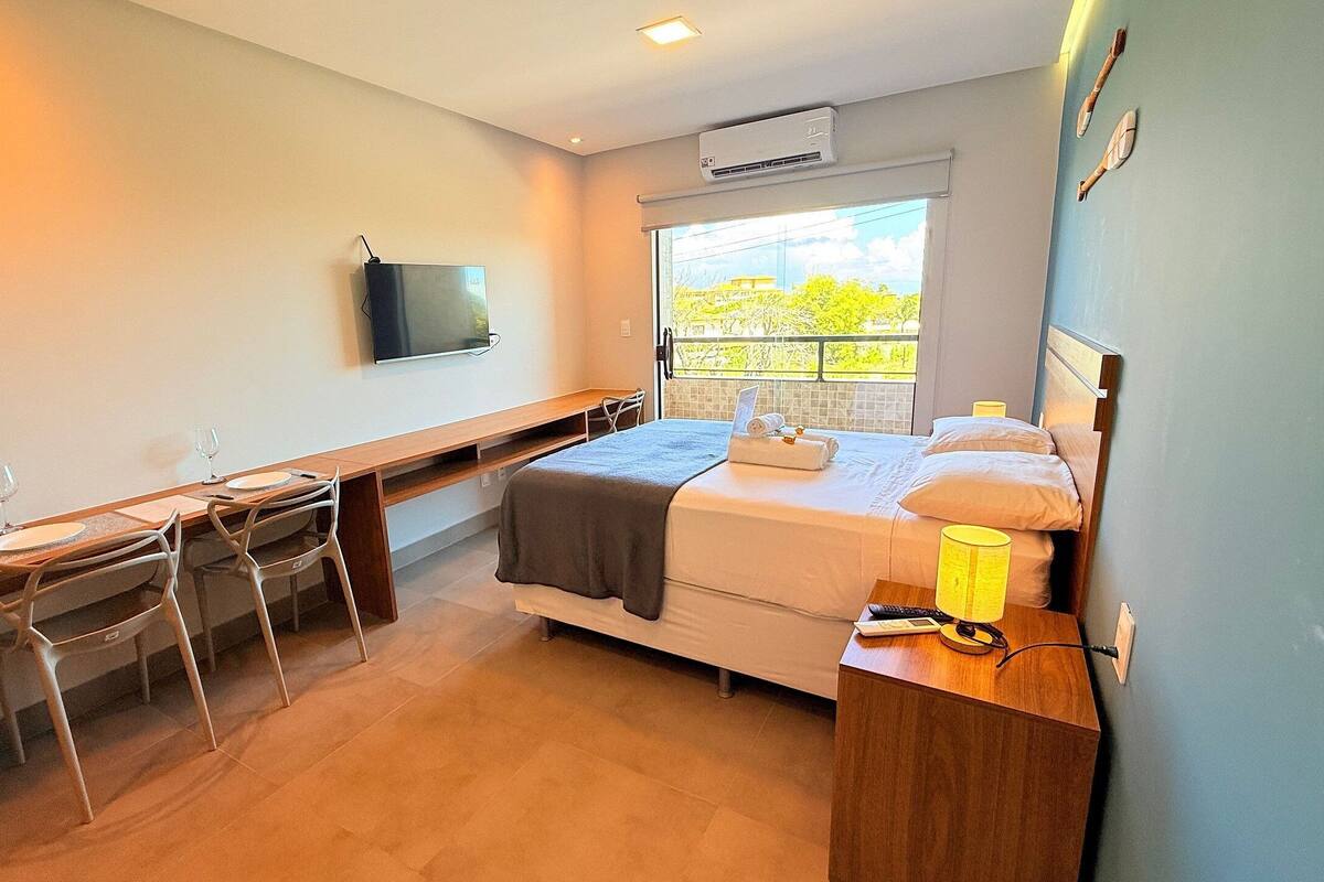 Quarto de hotel pequeno com cama, mesa de cabeceira, escrivaninha, cadeiras, televisão e janela com vista.