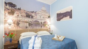 Quarto de hotel com cama, mesa de cabeceira, luminária, vaso com flores, bandeja com amenities e um mural na parede retratando uma paisagem urbana.