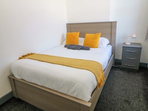Uma cama bem arrumada com cabeceira, uma mesa de cabeceira com abajur e uma pequena cômoda.