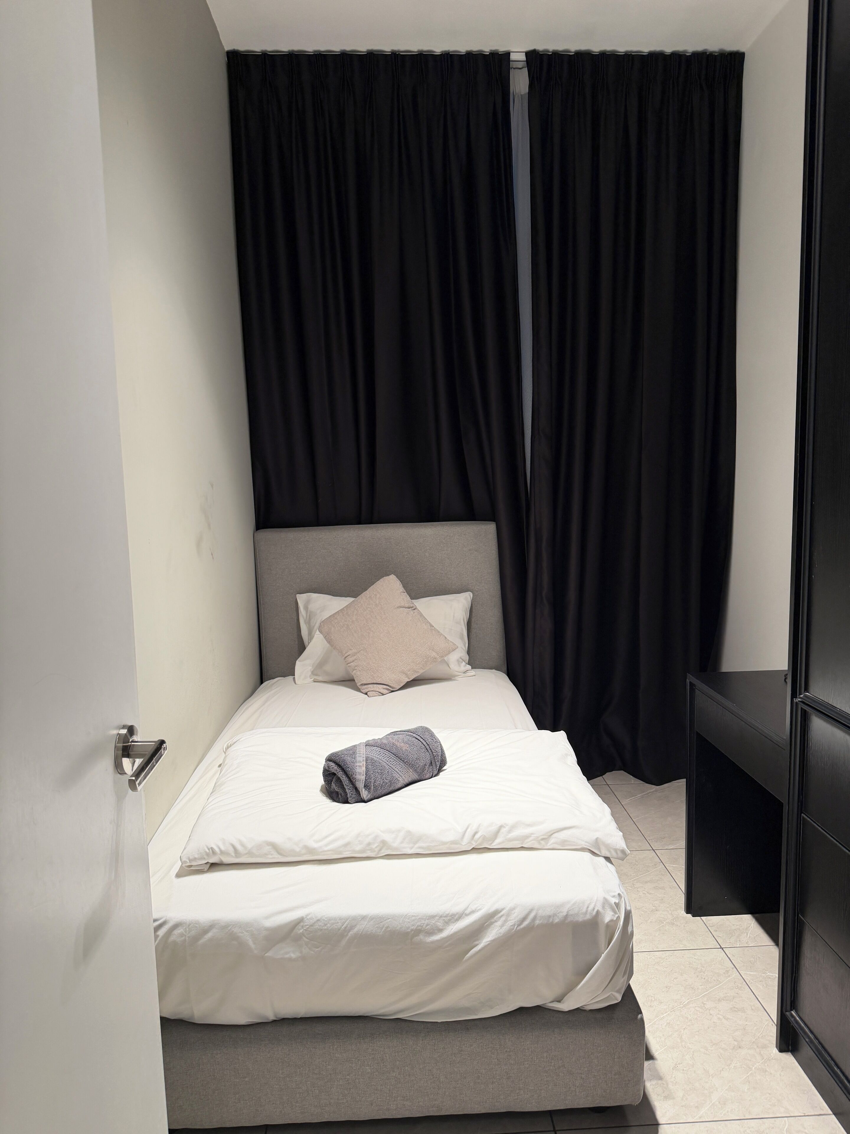 Uma cama pequena e bem arrumada, com cabeceira cinza, travesseiro bege e toalha cinza enrolada sobre a cama.