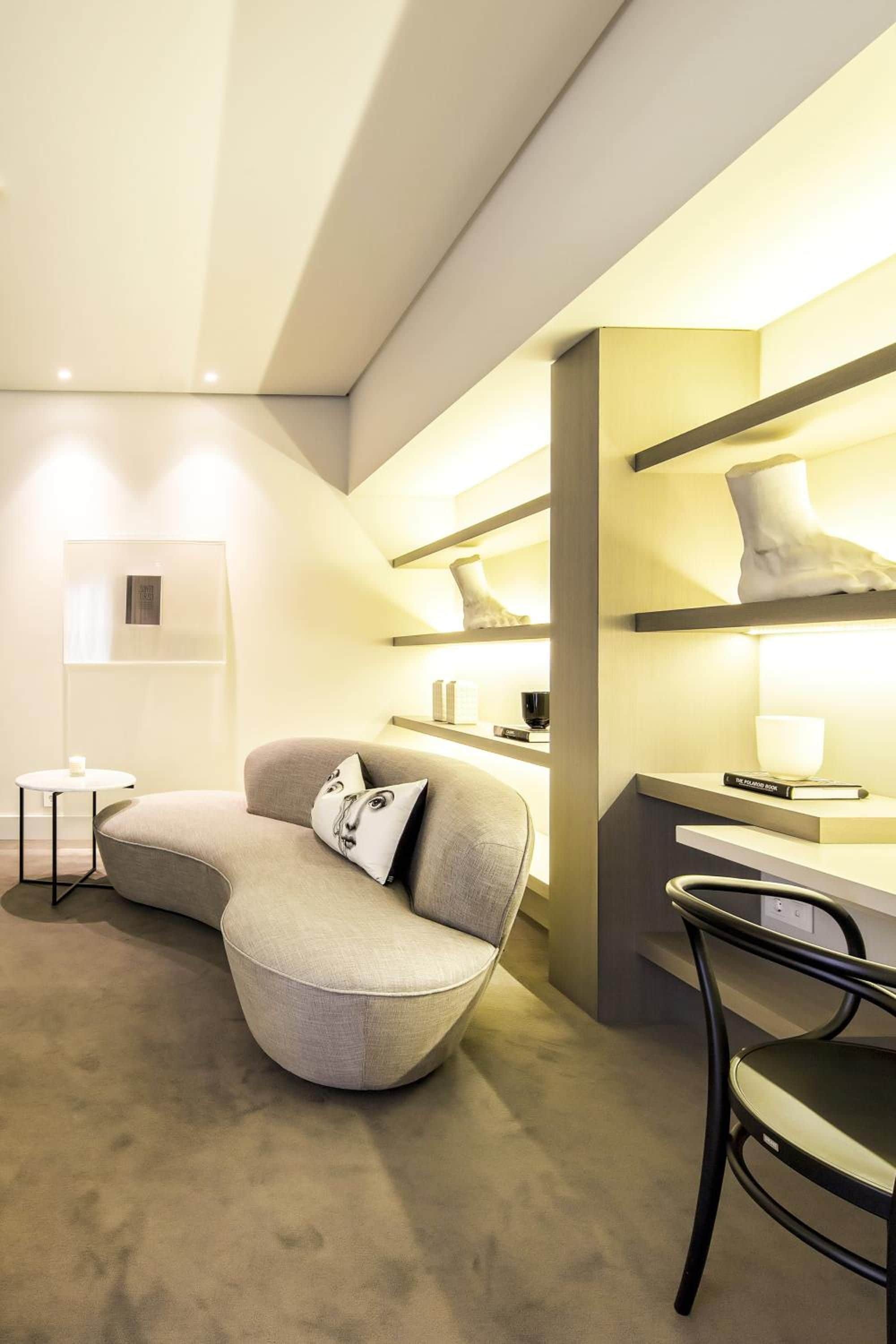 Um interior moderno com um sofá curvo, uma mesa redonda pequena e uma cadeira. Prateleiras exibem livros e objetos decorativos.