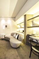 Um interior moderno com um sofá curvo, uma mesa redonda pequena e uma cadeira. Prateleiras exibem livros e objetos decorativos.