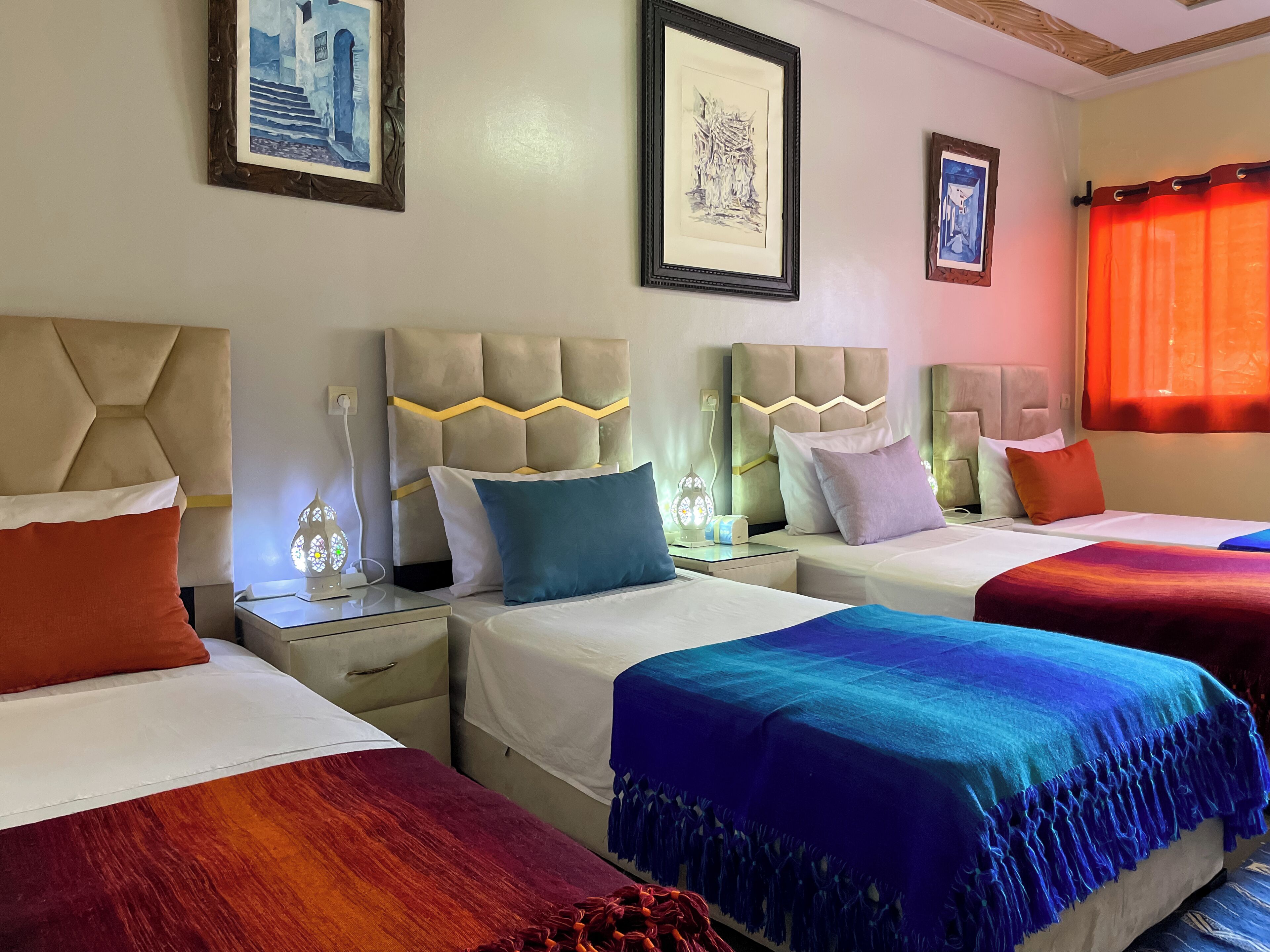 Quarto de hotel com três camas, cada uma com colcha de cor diferente, mesa de cabeceira com luminária e quadros nas paredes.