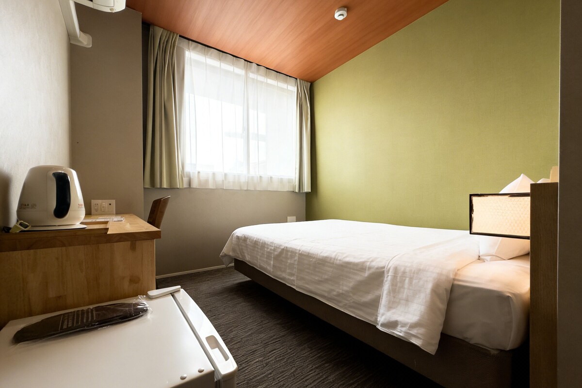 Quarto de hotel com uma cama, uma escrivaninha com chaleira, uma cadeira e uma janela com cortinas.