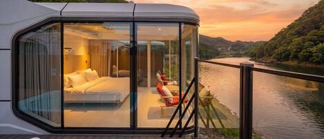 Um quarto moderno, com paredes de vidro, possui uma cama, uma poltrona vermelha e vista para um rio ao pôr do sol.