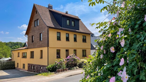 Uma casa de dois andares com telhado inclinado, várias janelas e um jardim com plantas floridas.