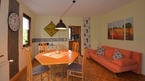 Uma sala de jantar com mesa de madeira, cadeiras e luminária pendente. Há um sofá e uma parede com quadros.