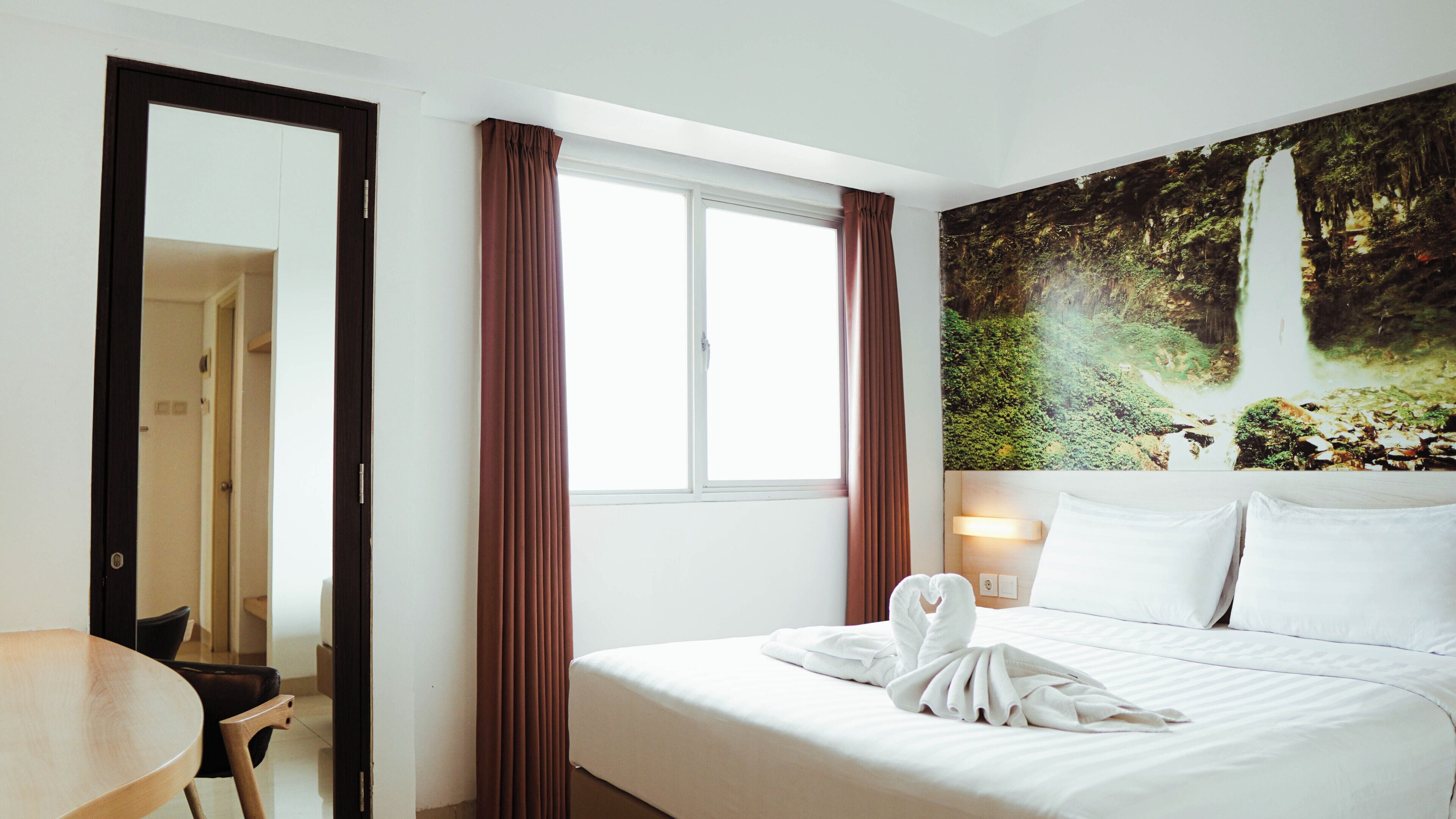Quarto de hotel com cama, espelho grande, janela com cortinas, pintura de uma cascata e mesa com cadeira.