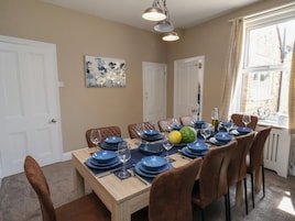 Uma sala de jantar com uma mesa de madeira posta para oito pessoas, cadeiras estofadas marrons e um quadro na parede.