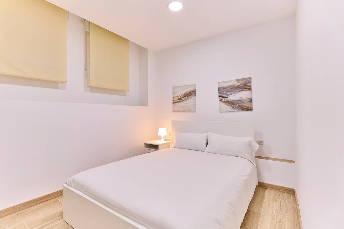 Quarto com cama, mesa de cabeceira, luminária na parede e duas pinturas abstratas.