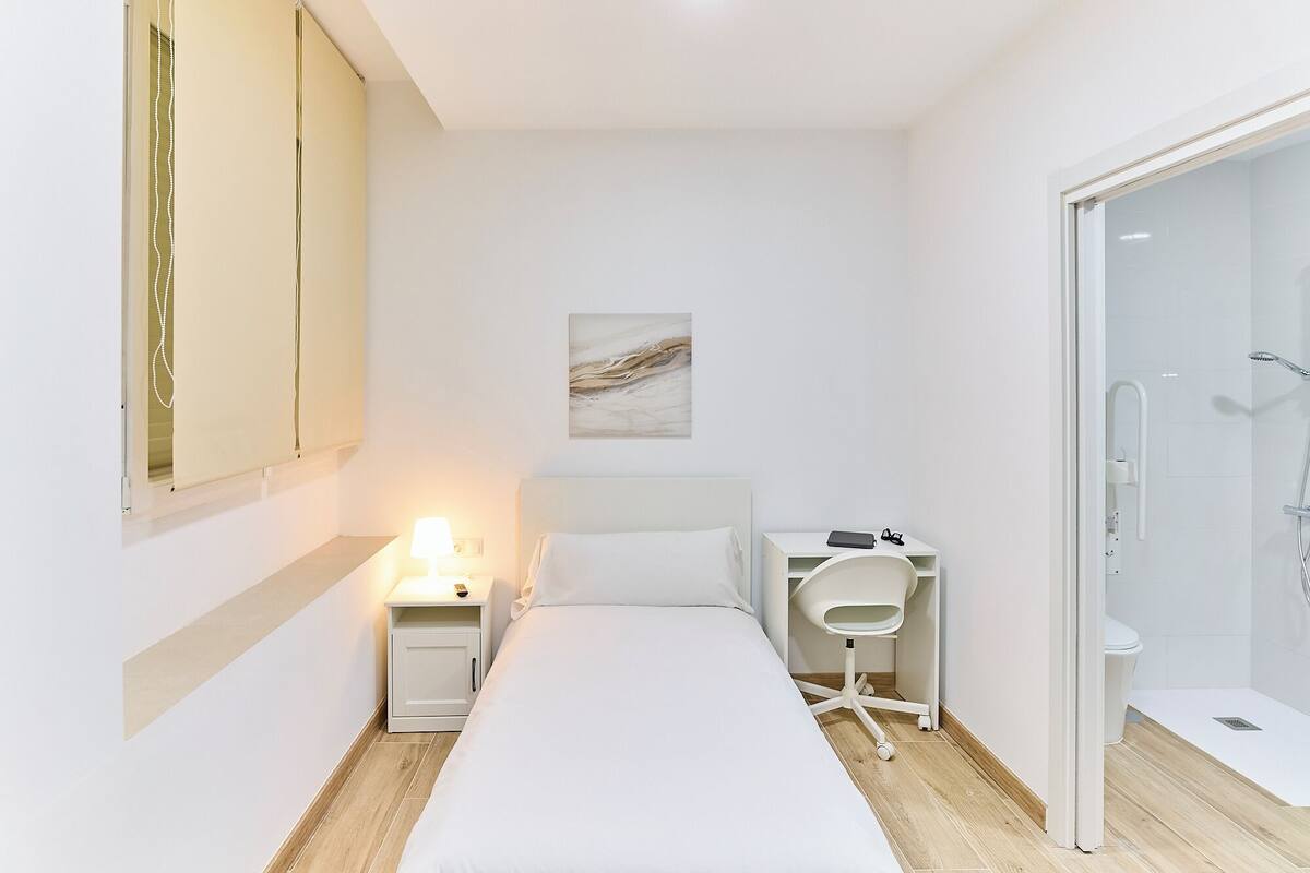 Quarto com cama, mesa de cabeceira, escrivaninha, cadeira e área de chuveiro.