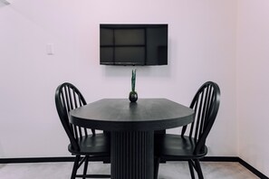 Uma mesa redonda com duas cadeiras pretas, uma televisão de tela plana fixada na parede acima e um pequeno vaso decorativo sobre a mesa.