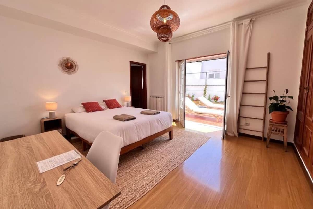 Quarto com cama, mesa de cabeceira, abajur e vista para área externa através de uma porta.