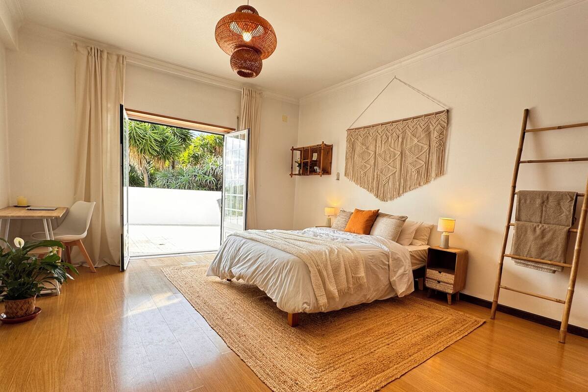 Quarto com cama, mesas de cabeceira, luminária pendente, escada e vista para o verde lá fora.