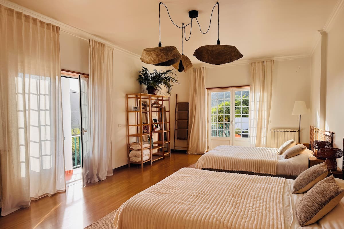 Quarto de hotel com duas camas, uma janela grande, uma planta e uma prateleira de madeira.