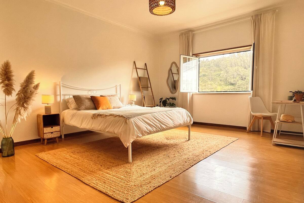Um quarto com cama, mesa de cabeceira, abajur, janela e cadeira.