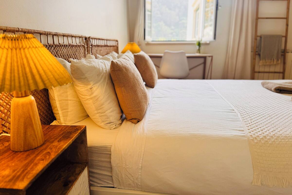 Uma cama bem arrumada com roupa de cama branca e cabeceira de madeira, uma mesa de cabeceira com abajur e uma janela com vista para prédios.