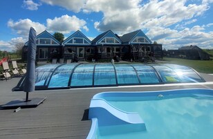 Piscina com estrutura de vidro, deck de madeira e um edifício com várias janelas e teto azul.