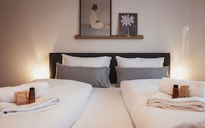 Quarto duplo com duas camas, cada uma com roupa de cama branca e almofadas cinzentas. Há toalhas e produtos de higiene pessoal nas camas.