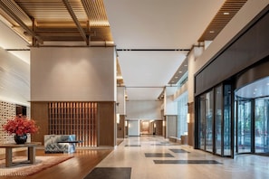 Um lobby moderno de hotel com um balcão de recepção central, piso estampado e uma grande porta de vidro que dá para o exterior.
