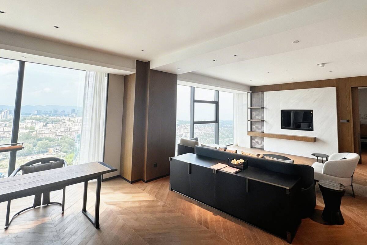 Uma sala de estar moderna com uma janela grande, um sofá preto, uma mesa de jantar e uma TV fixada na parede.
