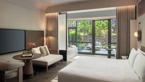 Quarto de hotel moderno com uma cama grande, área de estar com uma mesa e varanda com vista para o verde.