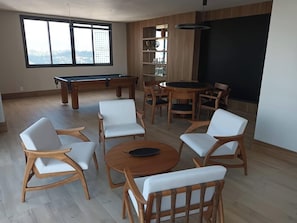 Uma sala de estar moderna com mesa de bilhar, área de jantar com cadeiras de madeira e vista da cidade por grandes janelas.