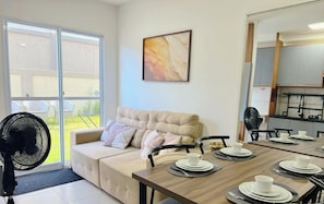 Uma sala de jantar com uma mesa de madeira posta para quatro pessoas, um sofá bege com almofadas, um ventilador preto e uma janela grande com vista para um jardim.