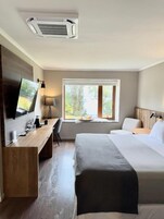 Quarto de hotel com cama, escrivaninha com cadeira, televisão e janela com vista para árvores.