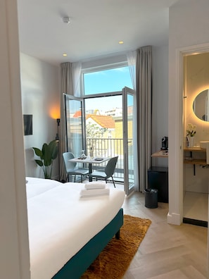 Quarto de hotel com cama, mesa de jantar com cadeiras, uma pequena varanda, banheiro com espelho e um cesto de lixo.