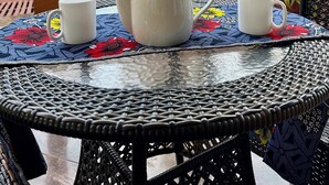 Uma mesa redonda com uma chaleira branca e duas chávenas brancas sobre uma toalha com padrão, rodeada por vegetação e um toldo branco.