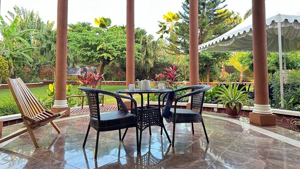 Um espaço ao ar livre coberto, com mesa e cadeiras, rodeado por vegetação e colunas.