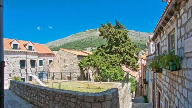 Uma rua de pedra estreita com edifícios de pedra tradicionais, um parquinho e uma colina ao fundo.